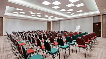 Sala de conferências com fileiras de cadeiras, um quadro branco e piso coberto de carpete.