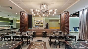 Um restaurante com várias mesas e cadeiras, área de bar e vista para o exterior.