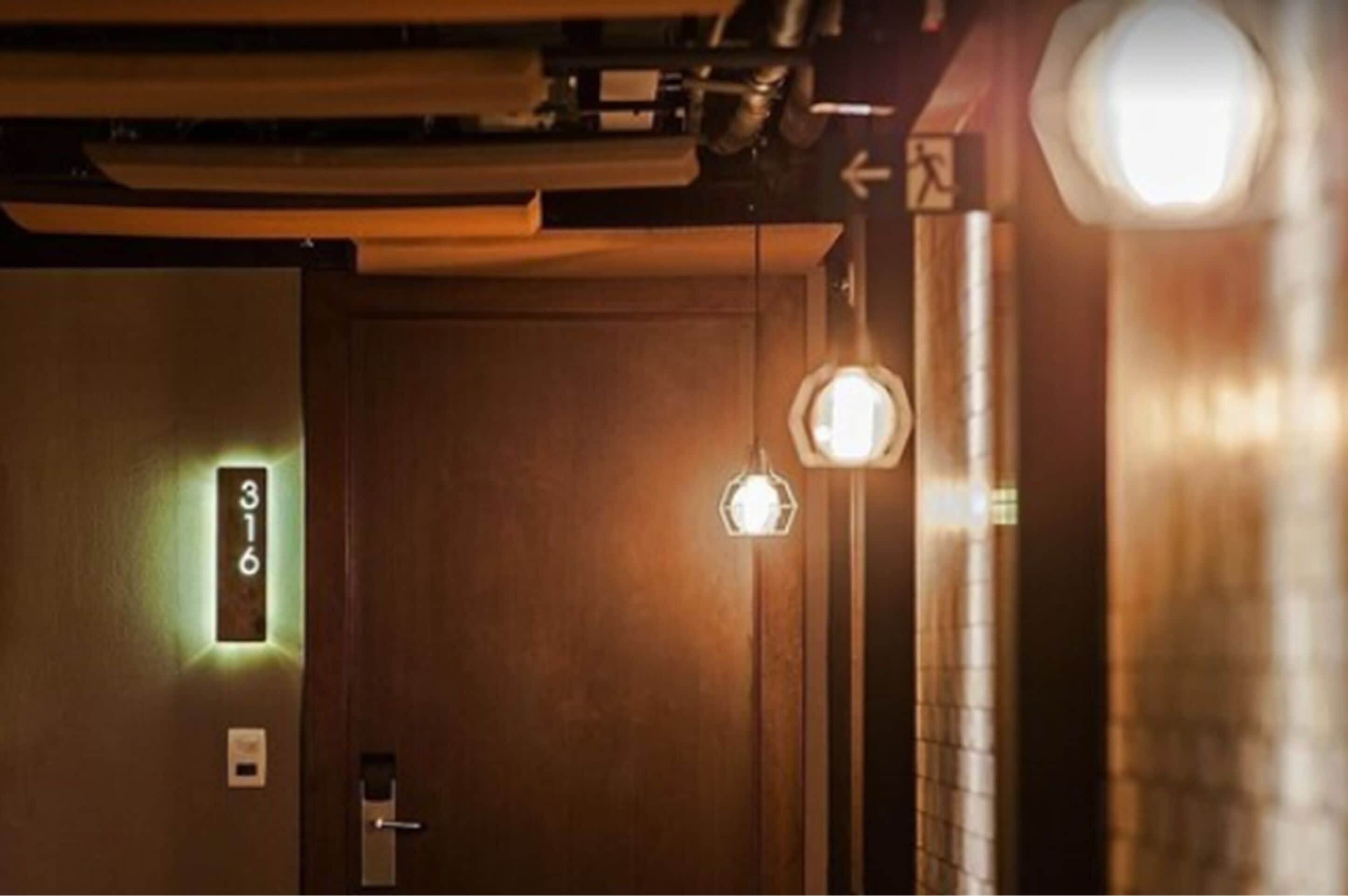 Quarto de hotel com porta de madeira, placa de identificação, luminária e luz embutida na parede.