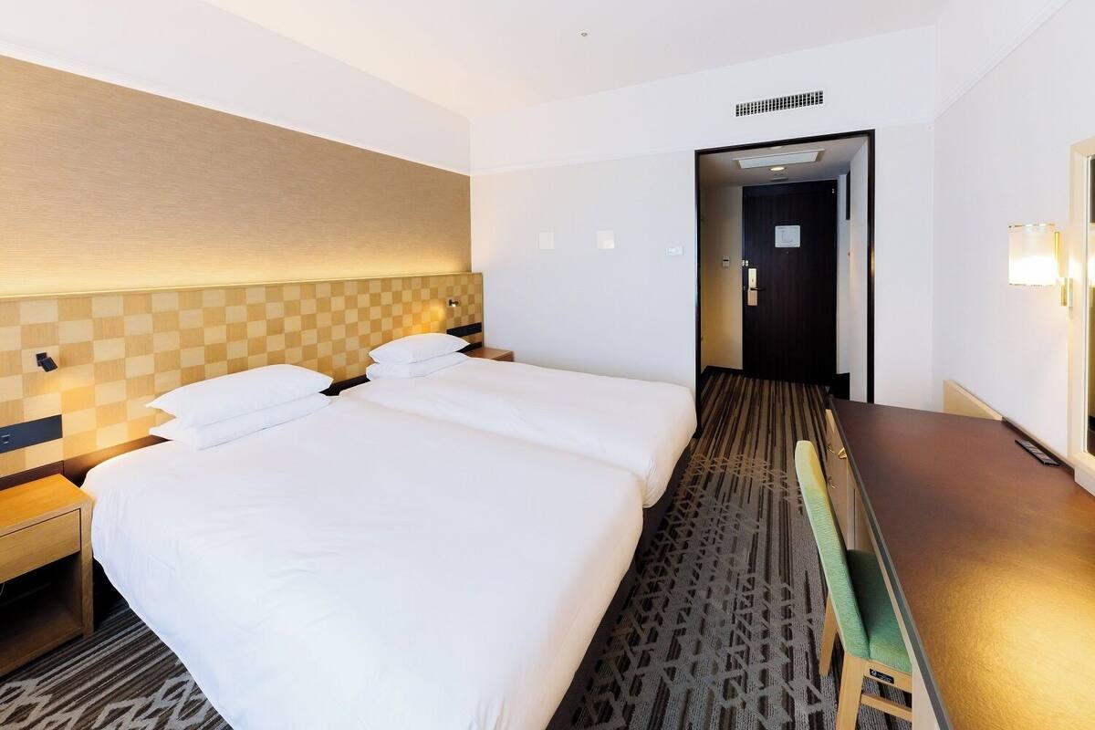Quarto de hotel com uma cama grande, uma escrivaninha e uma cadeira. Há uma porta que dá para outro quarto.
