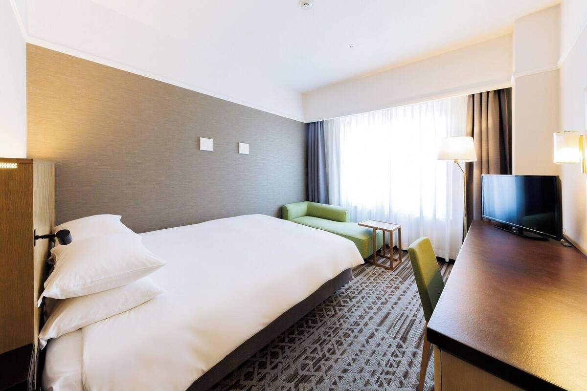 Quarto de hotel com uma cama grande, uma escrivaninha com televisão, uma mesinha e uma cadeira verde.