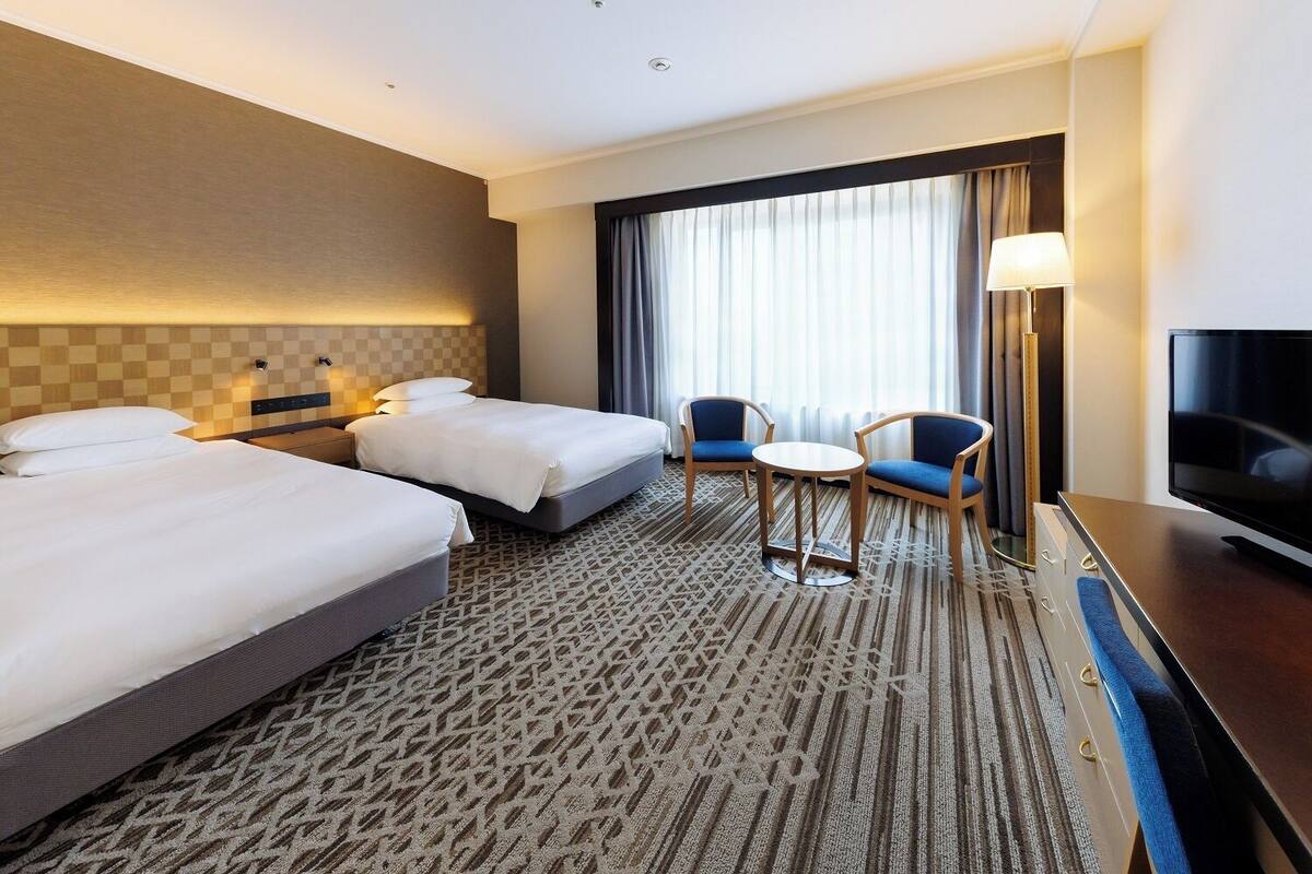 Quarto de hotel com duas camas, televisão, mesa pequena, duas cadeiras e uma janela grande com cortinas.