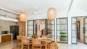 Ampla sala de estar com uma mesa de jantar posta para quatro pessoas, cercada por cadeiras de vime. Um ventilador de teto e luminárias pendentes de tecido são visíveis.