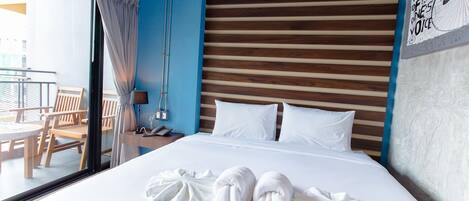 Quarto de hotel com cabeceira de madeira, cama com roupa de cama branca, duas toalhas sobre a cama, mesa de cabeceira com abajur e telefone, e vista para a varanda.