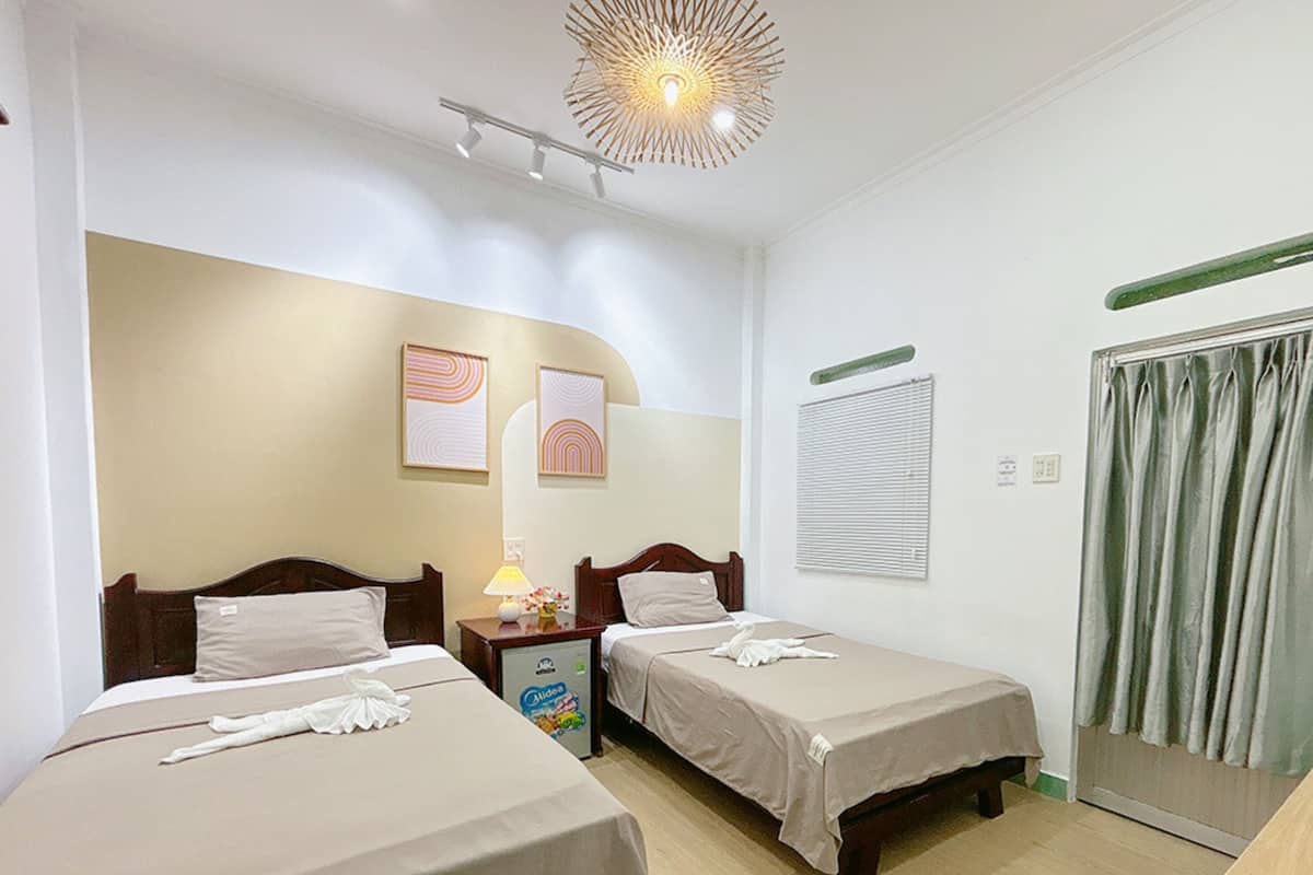 Quarto de hotel com duas camas de solteiro, uma mesa de cabeceira com abajur, um mini-refrigerador e uma janela com persianas.