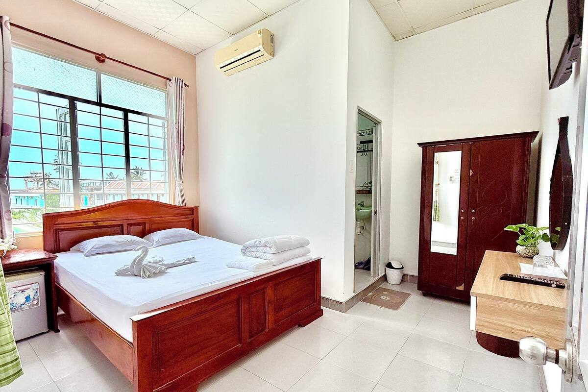 Quarto de hotel com cama de madeira, escrivaninha, guarda-roupa e banheiro visível por uma porta aberta.