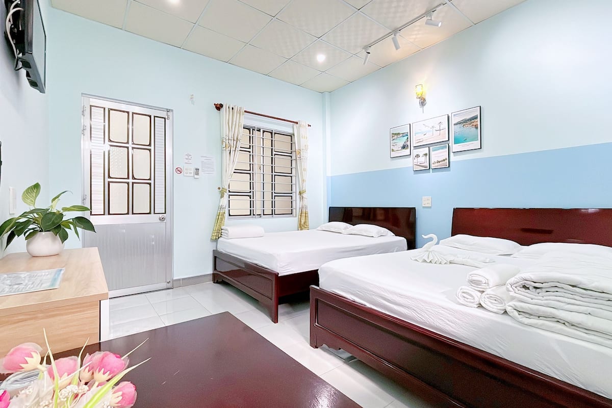 Quarto de hotel com duas camas, uma escrivaninha, um espelho, uma planta em vaso e uma janela com cortinas.