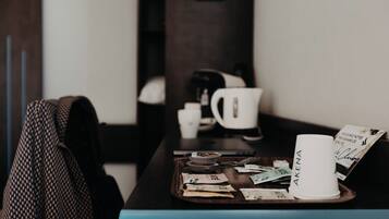 Quarto de hotel com uma mesa de madeira, uma caneca branca com a inscrição 'AKENA', um tabuleiro com diversos objetos e uma bolsa com padrão preto e branco.
