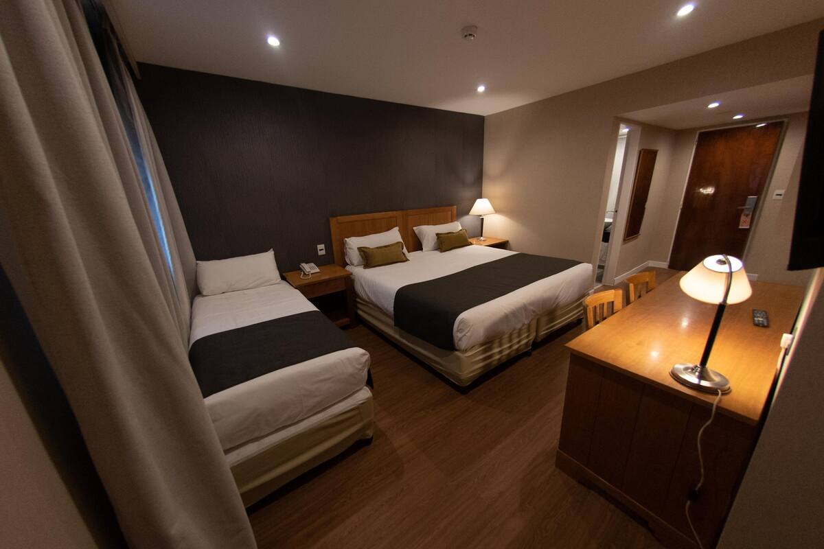 Quarto de hotel com duas camas, uma escrivaninha, uma cadeira, um abajur e uma televisão.