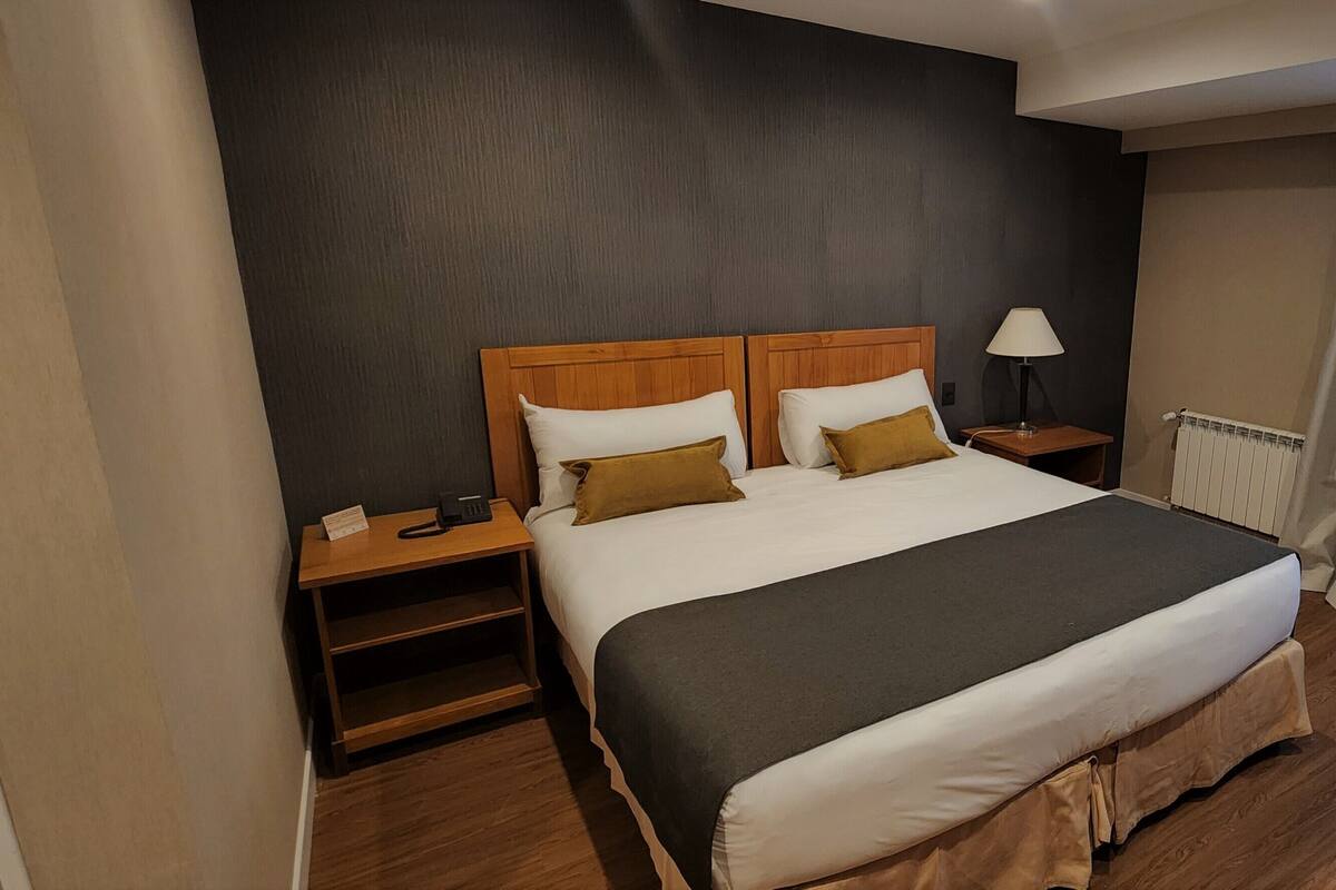 Quarto de hotel com uma cama grande, cabeceira de madeira, duas mesas de cabeceira, um abajur, um radiador e uma janela com cortinas.