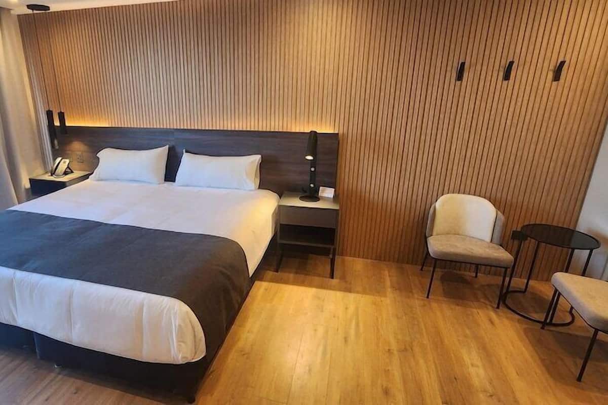 Quarto de hotel com uma cama grande, mesas de cabeceira, uma cadeira e uma pequena mesa.