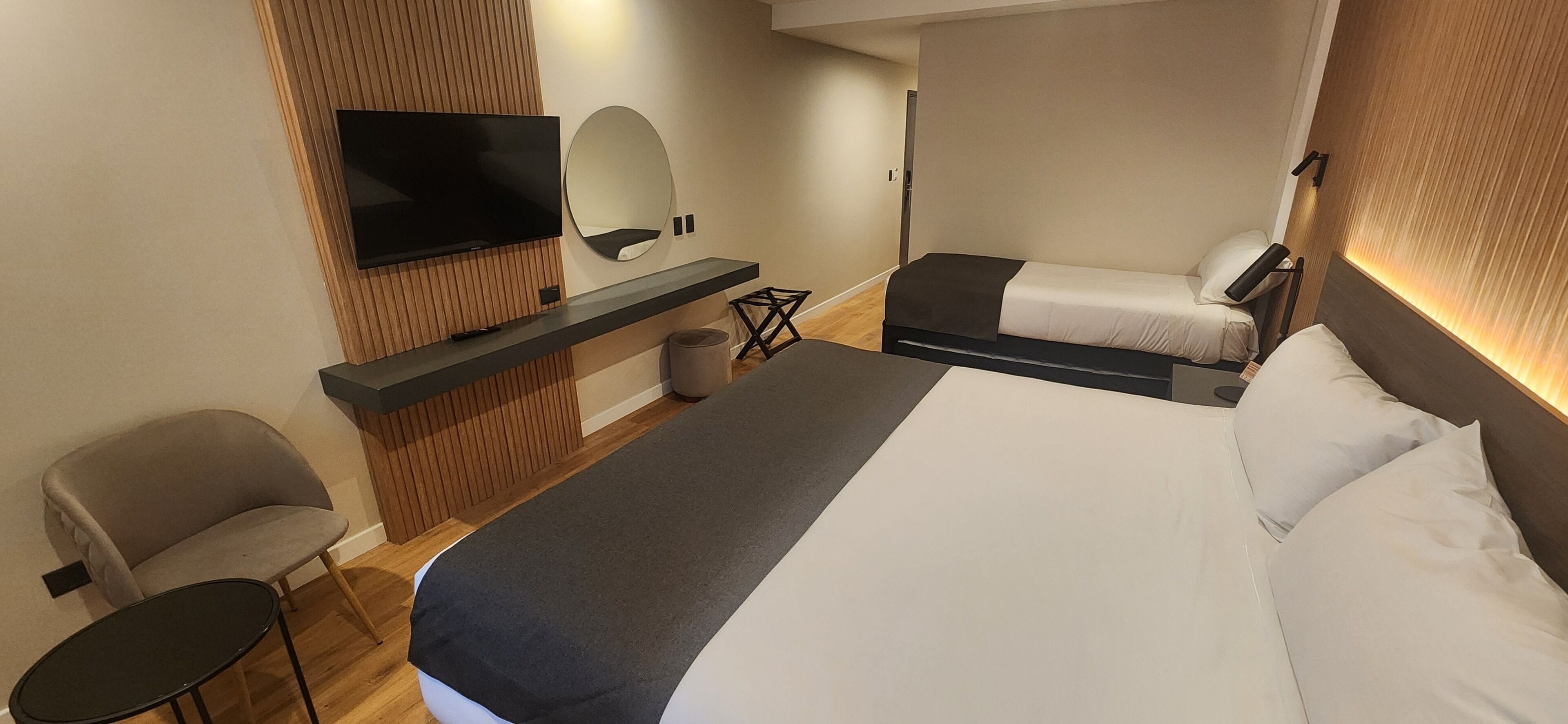 Quarto de hotel com uma cama grande, uma televisão na parede, uma mesinha de cabeceira, uma cadeira e uma pequena mesa.