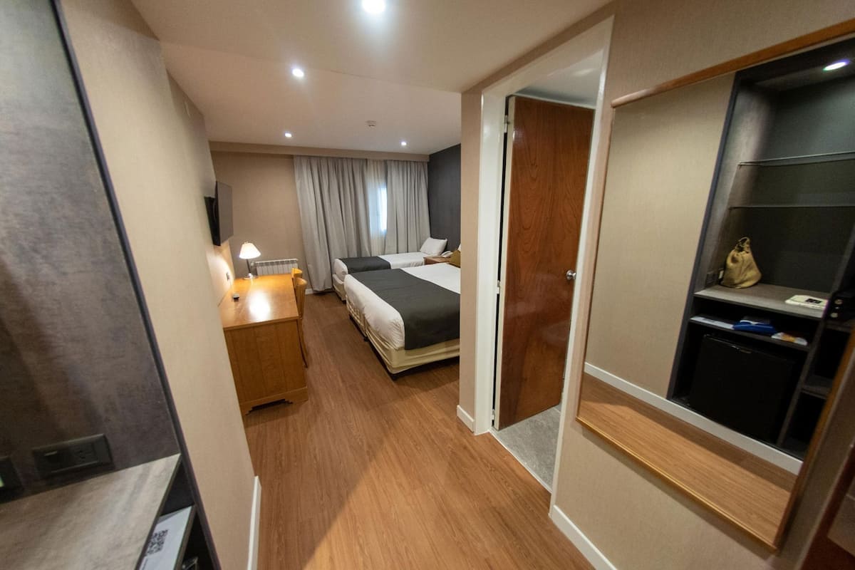 Quarto de hotel com cama, mesa de cabeceira, luminária, guarda-roupa e pequena escrivaninha com cadeira.