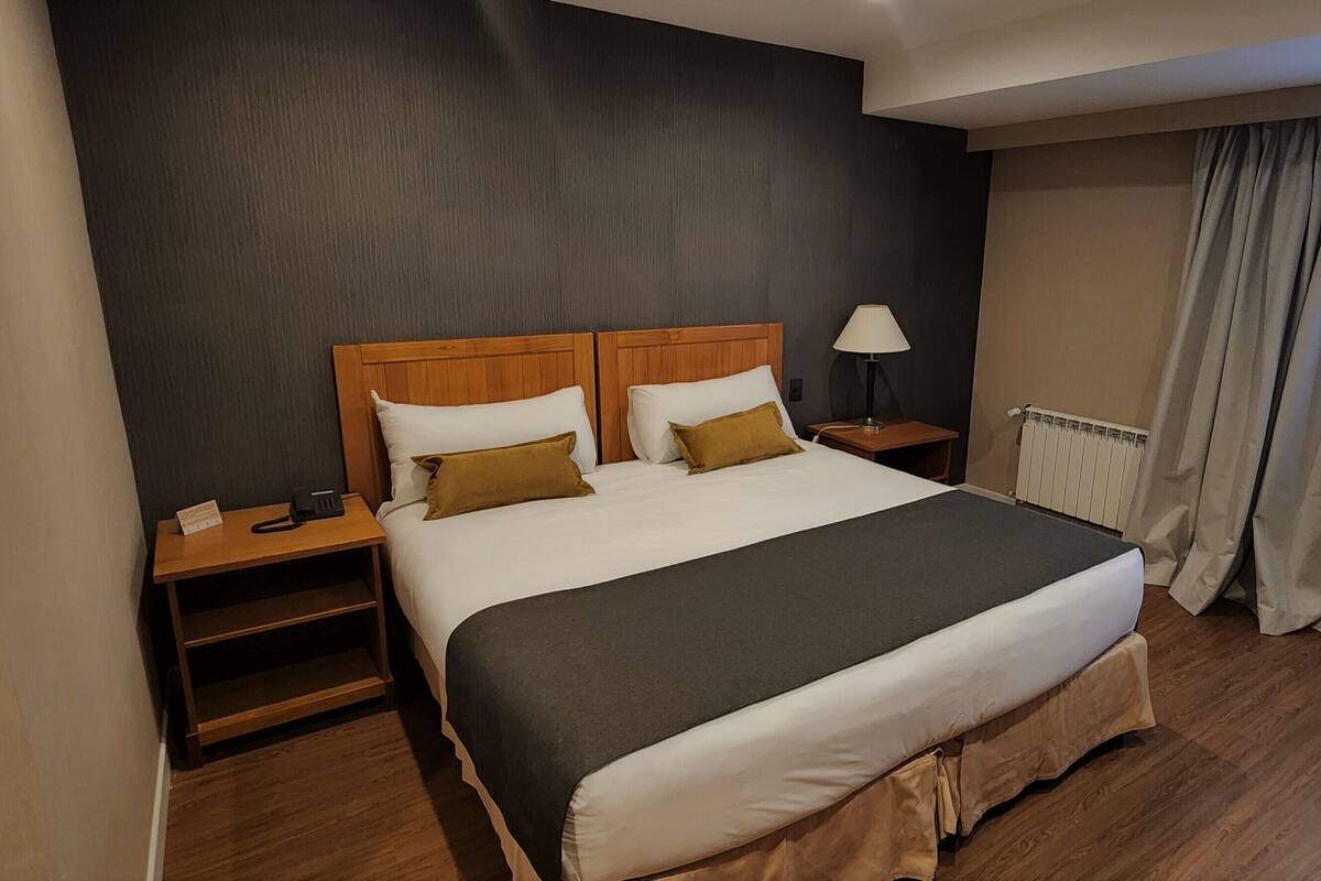 Quarto de hotel com cama grande, cabeceira de madeira, mesas de cabeceira, luminária, radiador e janela com cortinas.
