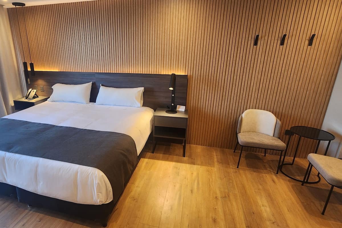 Quarto de hotel com uma cama grande, mesas de cabeceira, uma cadeira e vista para o exterior.