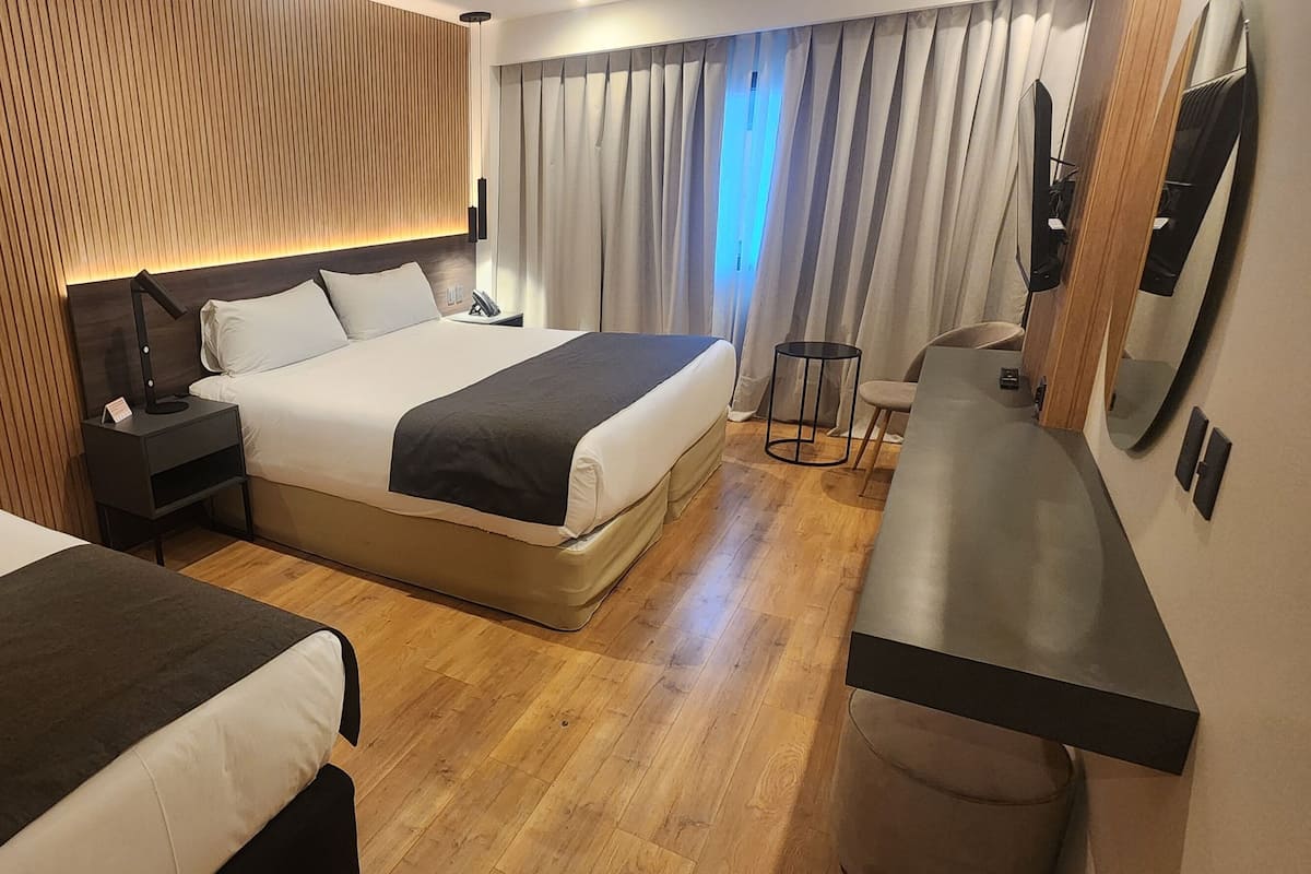 Quarto de hotel com duas camas, uma escrivaninha, uma cadeira, um espelho e teto de madeira.