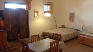 Um quarto pequeno e acolhedor, com uma cama, uma mesa com cadeiras, uma escrivaninha e um abajur.