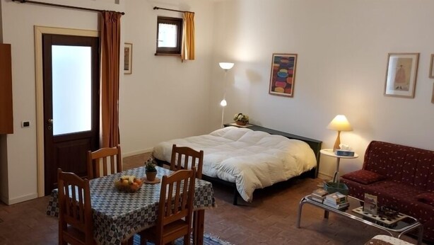 Quarto compacto com cama, mesa de jantar com cadeiras, sofá, pequena mesa com abajur e cozinha compacta.