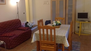 Uma sala de estar com um sofá vermelho, uma mesa de jantar com cadeiras, uma TV sobre um móvel e um armário com cestos.