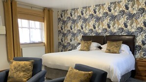 Quarto de hotel com uma cama grande, duas poltronas, uma mesinha de cabeceira e uma janela com cortinas.