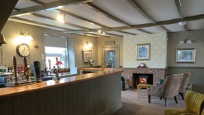 Uma área de bar com balcão de madeira, torneiras de cerveja e um relógio. Há uma lareira com parapeito e quadros emoldurados. Dois cadeiras estão visíveis na área de estar.