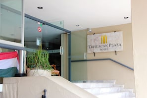 Entrada de hotel com placa indicando Tehuacan Plaza Hotel, porta de vidro, planta em vaso e uma bandeira.