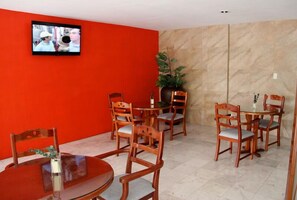 Uma sala de jantar com cadeiras de madeira e uma mesa de vidro, uma televisão fixada na parede e um vaso com planta.