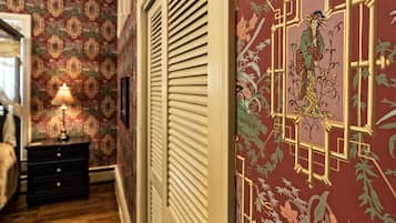Um corredor com papel de parede estampado, piso de madeira, mesa de cabeceira com abajur e um painel decorativo na parede.