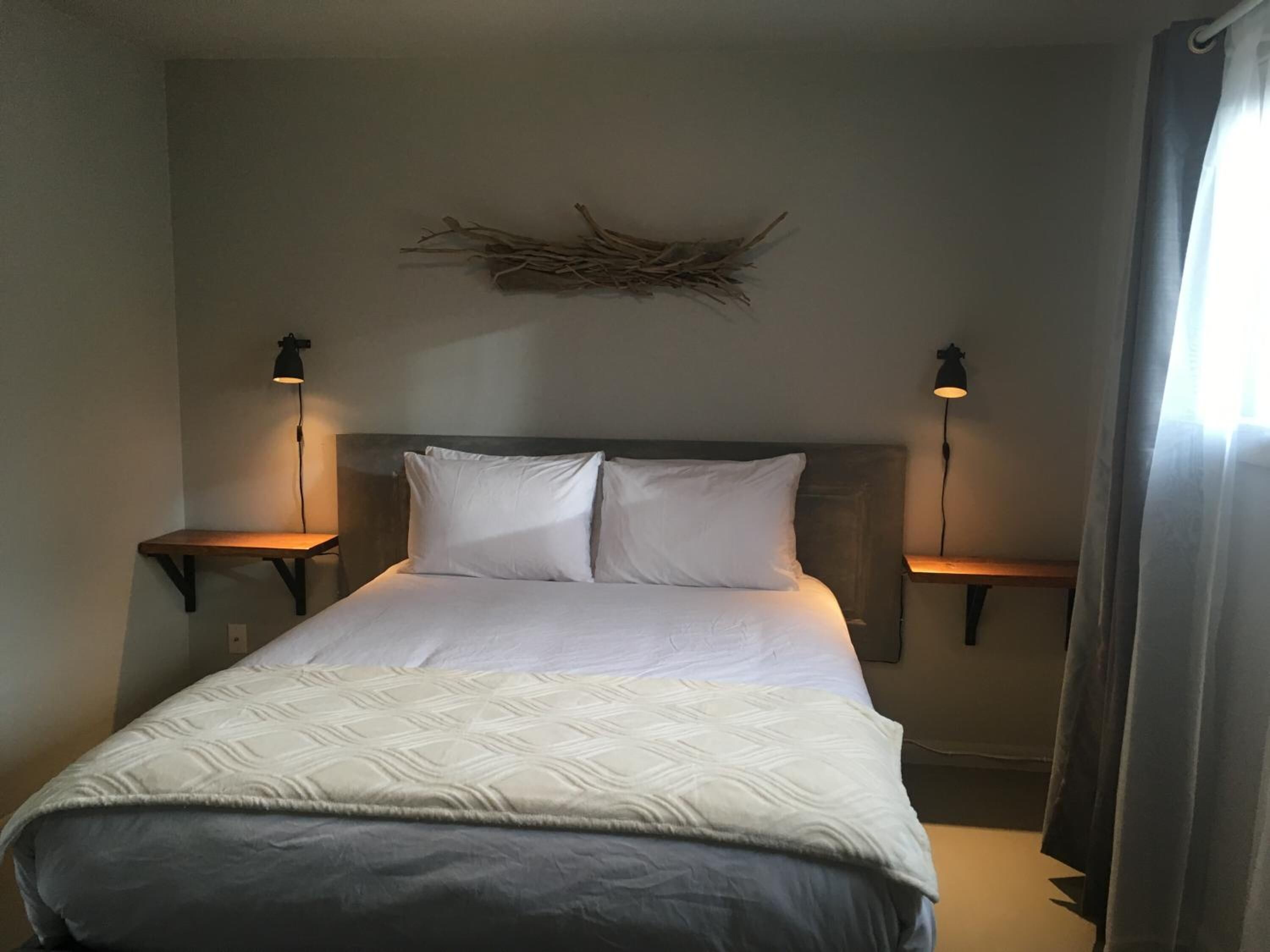 Uma cama bem arrumada com cabeceira, duas mesas de cabeceira com abajures e luminária na parede.