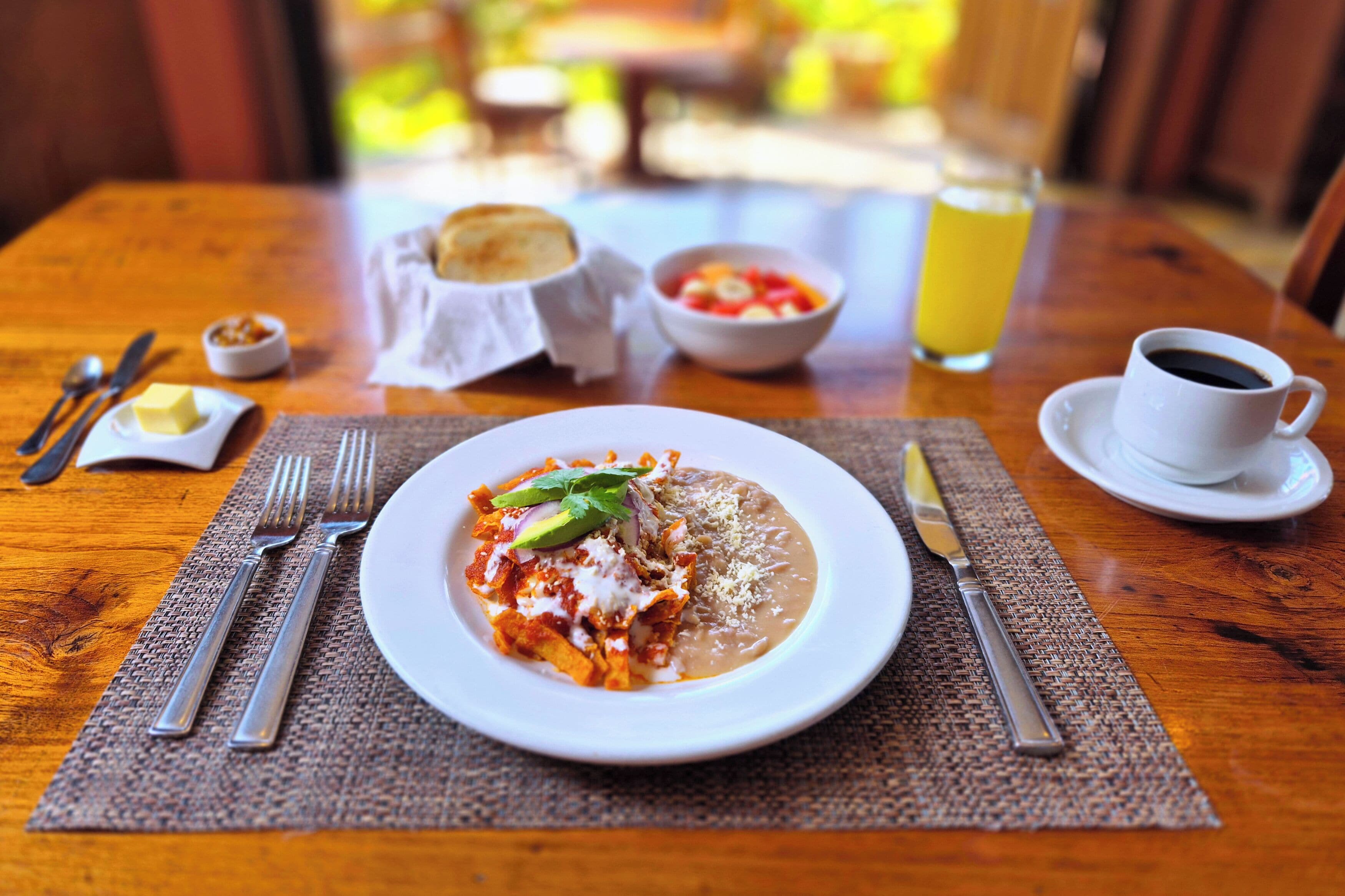 Uma mesa de jantar com um prato de enchiladas, uma tigela de fruta, um copo de suco de laranja e uma xícara de café.