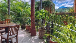 Uma área de pátio com uma mesa e cadeiras de madeira redondas, cercada por plantas em vasos e uma estátua.