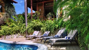 Uma área de piscina com espreguiçadeiras e banheira de hidromassagem, cercada por vegetação exuberante e uma parede de pedra.