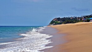 Uma praia arenosa com ondas suaves, céu claro e área residencial com casas e palmeiras ao fundo.