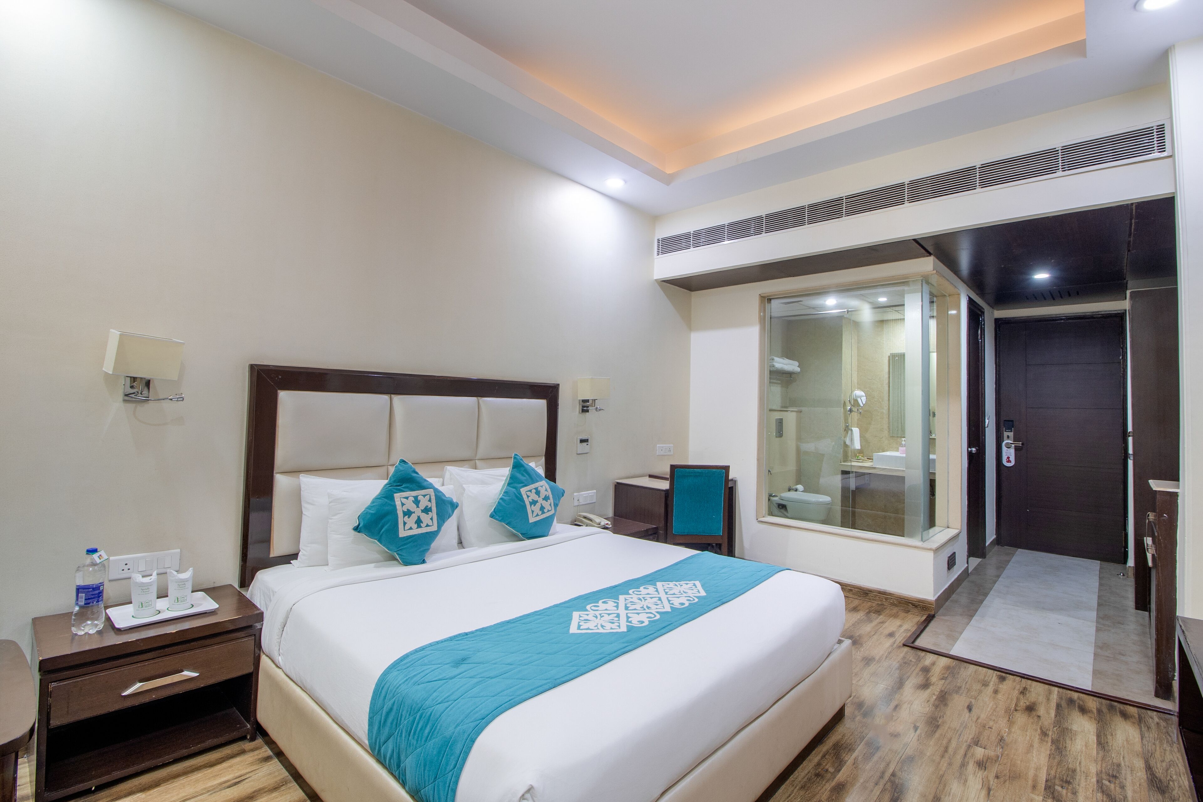 Quarto de hotel com uma cama grande, uma mesa de cabeceira com luminária, um criado-mudo, um banheiro com box de vidro e uma porta que dá para outro cômodo.