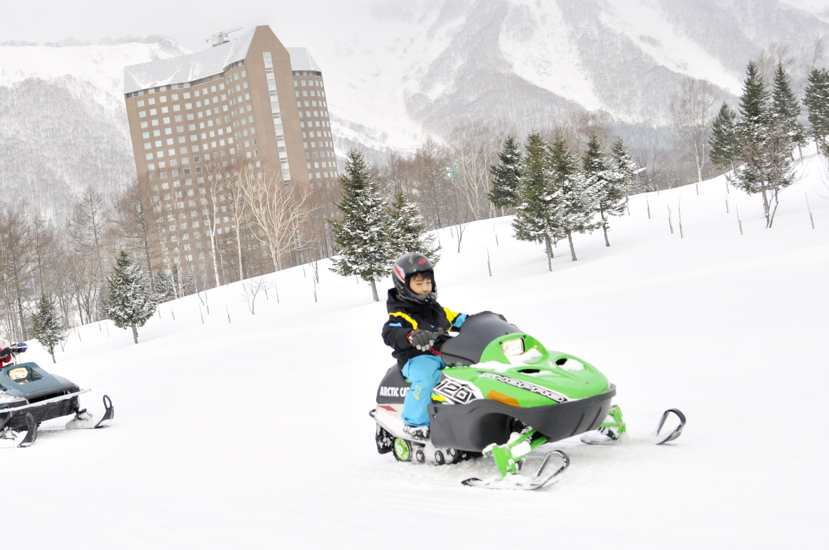 Uma moto de neve com um condutor num casaco preto e amarelo, uma moto de neve verde e preta, e um edifício ao fundo.