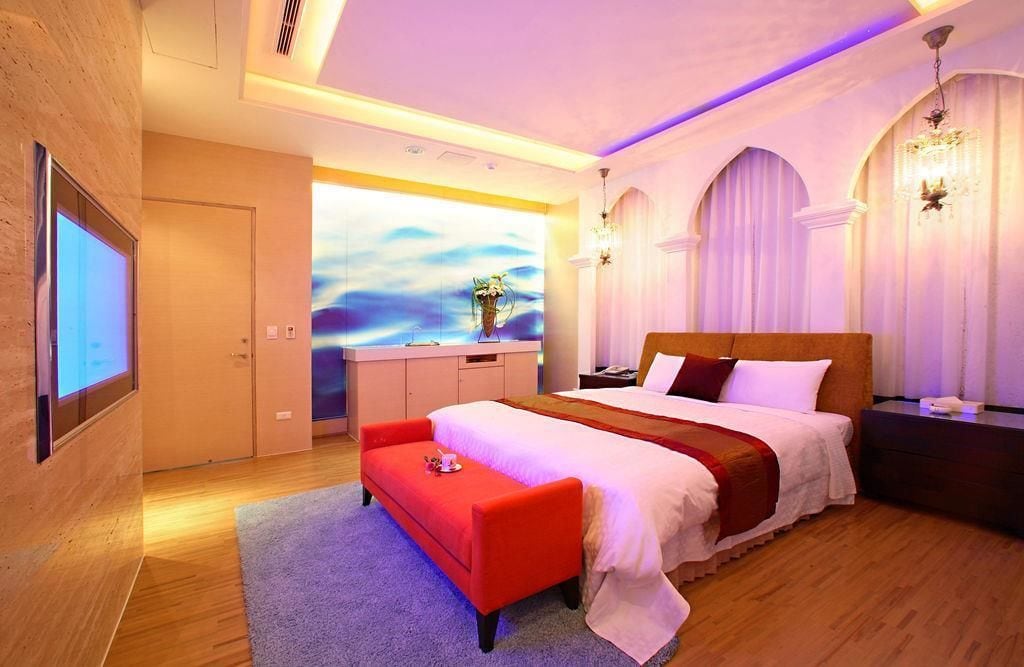 Quarto moderno com uma cama grande, um banco vermelho, uma toucador com espelho e uma TV fixada na parede.