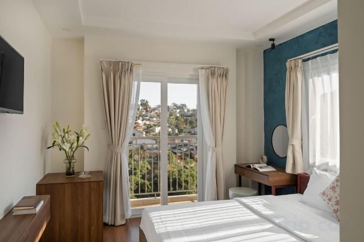 Quarto com cama, escrivaninha, cadeira, televisão e vista da cidade pela janela.