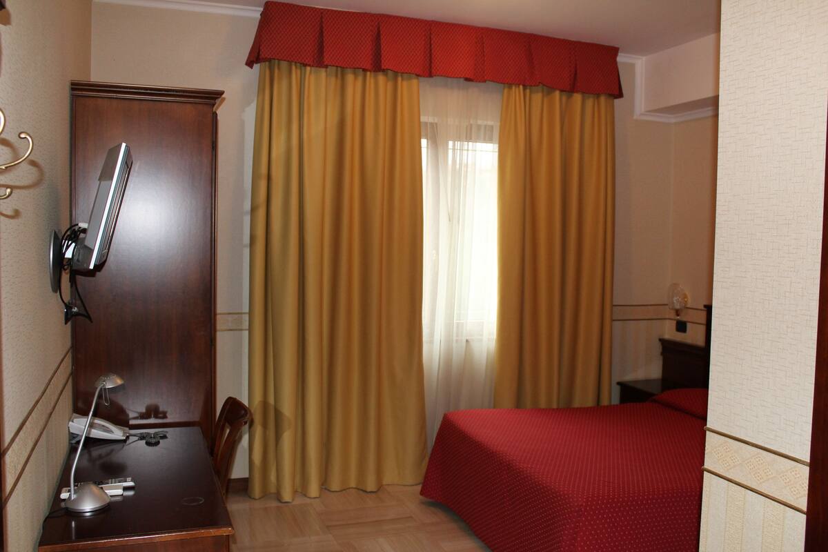 Quarto de hotel com escrivaninha de madeira, telefone, luminária, cama com colcha vermelha e janela com cortinas amarelas.