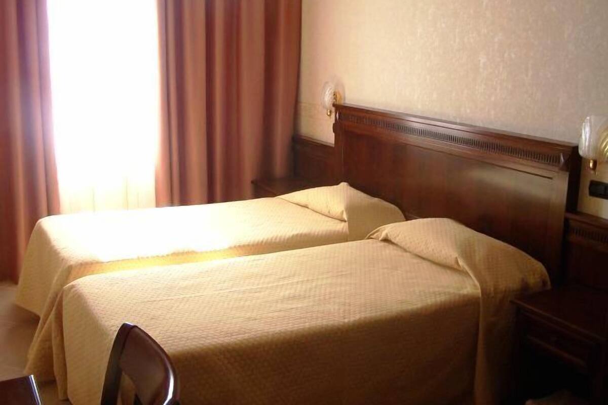 Quarto de hotel com duas camas, cabeceira de madeira, mesa de cabeceira e uma cadeira.