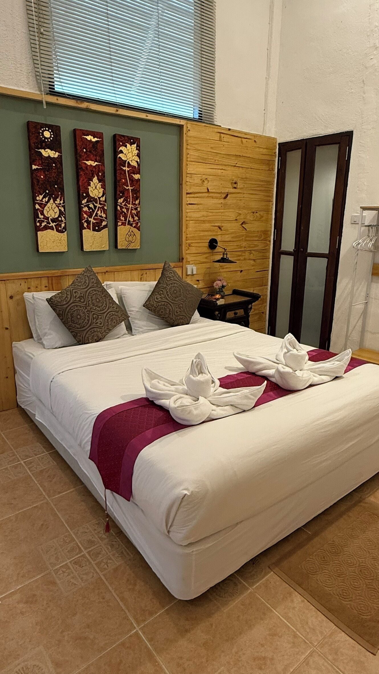 Cama bem arrumada com lençóis brancos e toalhas decorativas, cabeceira de madeira, mesa de cabeceira com abajur e três quadros na parede.
