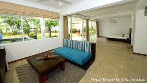 Ampla sala de estar com sofá azul, mesa de centro de madeira e ventoinha no teto. Janelas amplas oferecem vista para o exterior.