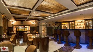 Uma área de bar com mesa de vidro e vários banquetes, um balcão de recepção e um bar abastecido com garrafas.