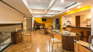 Um hall de hotel moderno com uma mesa de centro de vidro, cadeiras de madeira, um balcão de recepção e uma televisão fixada na parede.