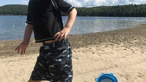 Criança brincando na areia de uma praia perto de um lago, com barcos e uma floresta ao fundo.