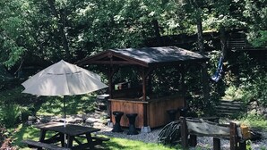 Um gazebo de madeira com uma mesa e um guarda-sol, cercado por árvores e um caminho de pedra.