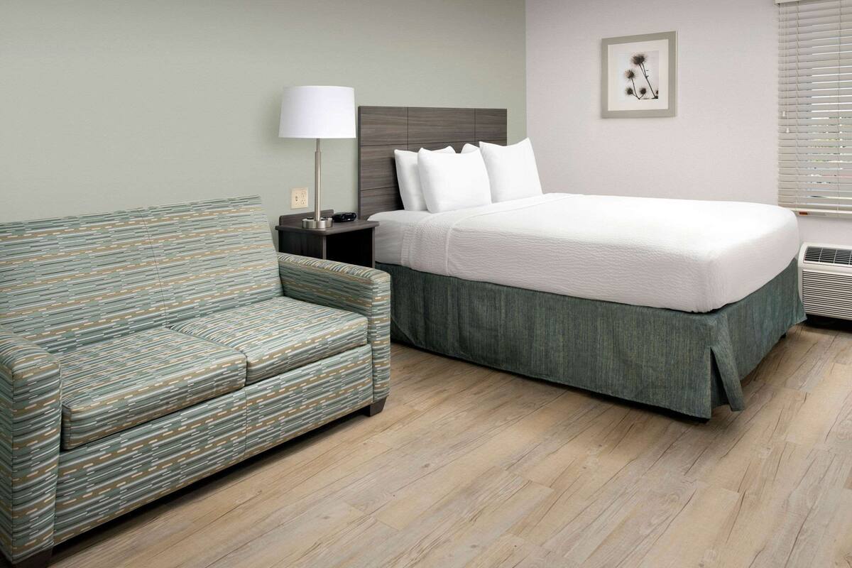 Quarto de hotel com cama, mesa de cabeceira, abajur, quadro na parede e janela com persianas.