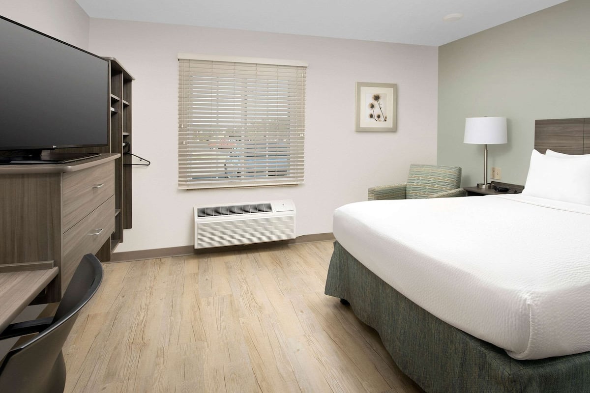 Quarto de hotel com cama, televisão, poltrona, luminária e quadro na parede.