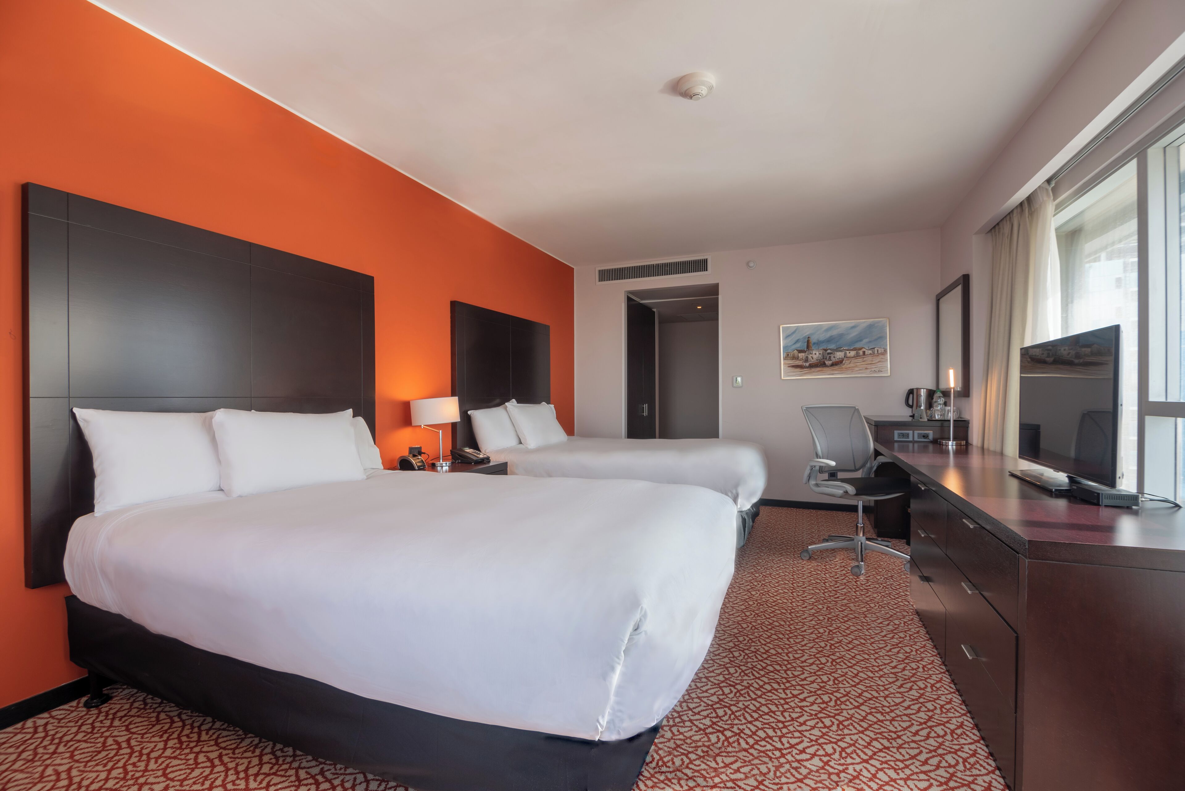 Quarto de hotel com duas camas, uma escrivaninha, uma cadeira e uma janela grande.