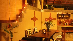 Uma área de refeições com uma mesa de madeira, cadeiras e um vaso com flores. Há decorações nas paredes e uma estante com objetos.