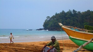 Uma praia com um homem sentado num tronco, um barco e uma mulher caminhando.